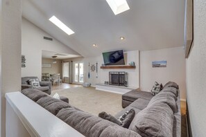 Uma sala de estar com um amplo sofá em formato de L, uma TV de tela plana na parede, uma lareira e uma área de jantar ao fundo.