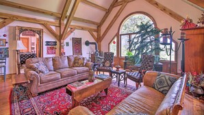 Uma sala de estar ampla com teto alto, vigas de madeira, uma grande janela com vista para árvores e diversos assentos, incluindo um sofá, poltronas e uma mesa de centro.
