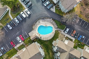 Vista aérea de uma área residencial com piscina, cercada por um deck de concreto, vagas de estacionamento próximas e casas com entradas de automóvel visíveis.