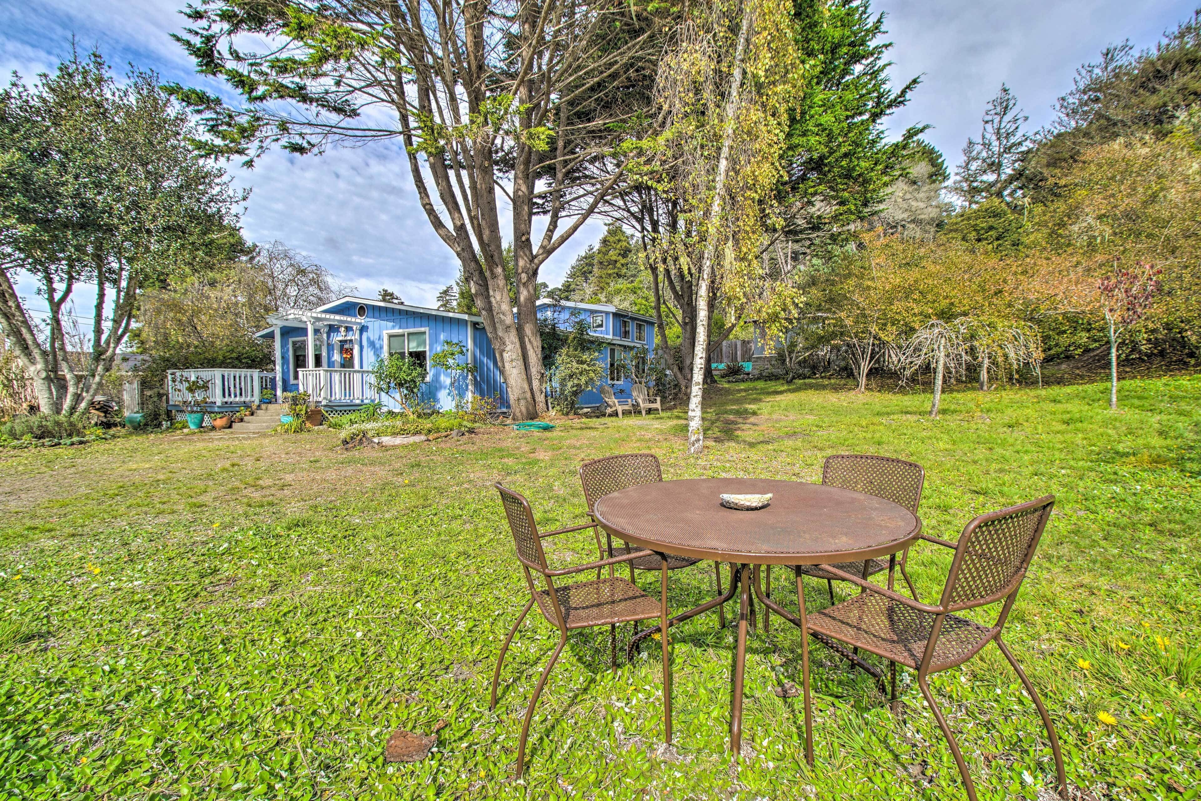 Uma casa azul com varanda, cercada por árvores e um gramado com uma mesa e cadeiras de metal.