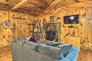 Interior acolhedor de uma cabana de madeira com lareira de pedra, paredes de madeira, uma televisão e uma área de estar confortável.