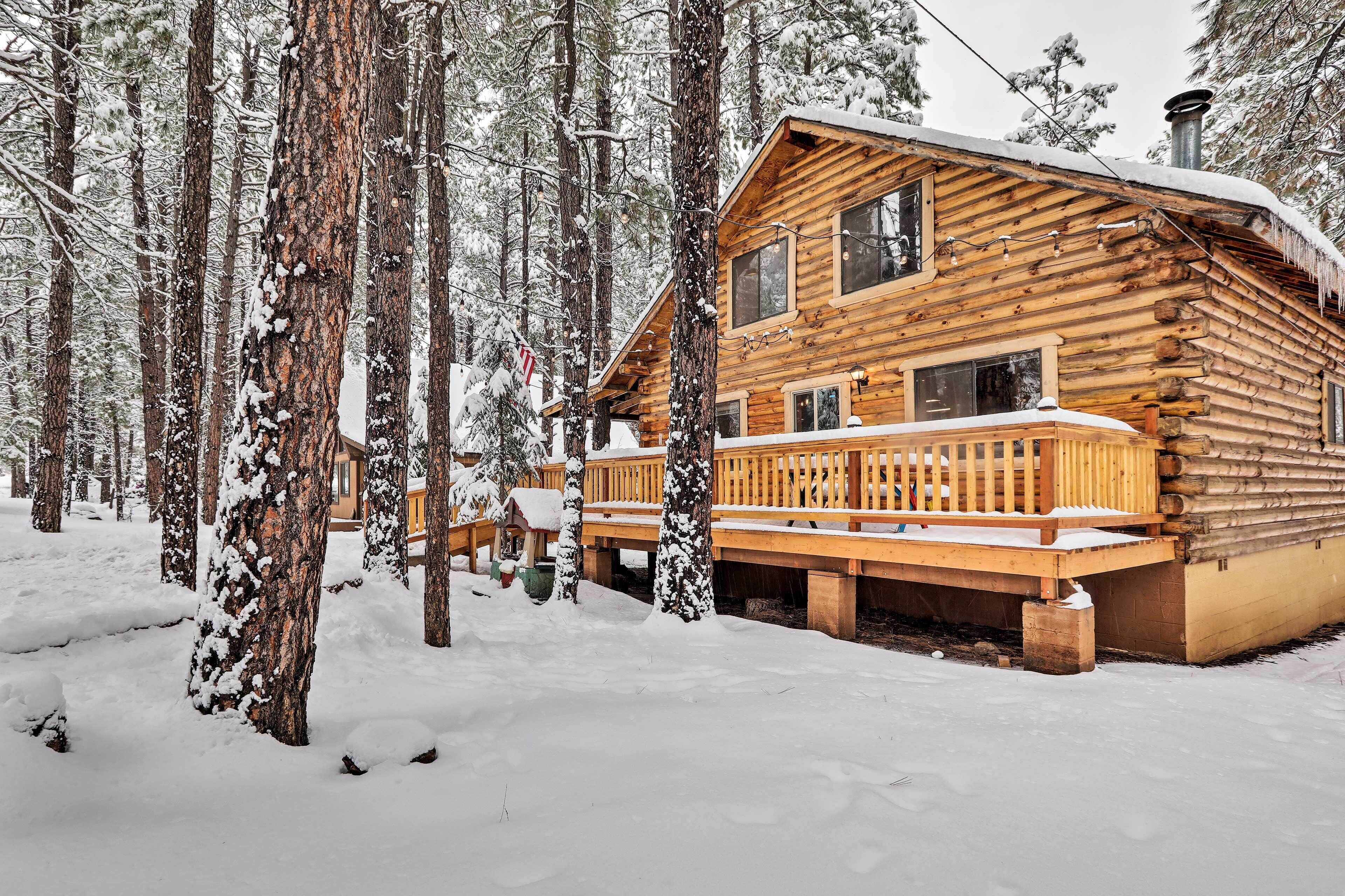 Uma cabana de troncos com deck de madeira e chaminé, cercada por árvores cobertas de neve.