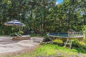 Uma área de piscina com espreguiçadeira e guarda-sol, rodeada por vegetação exuberante e um deck de madeira.