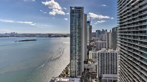 Um edifício alto com design arquitetônico moderno, localizado ao lado de um corpo d'água, com um horizonte urbano ao fundo.