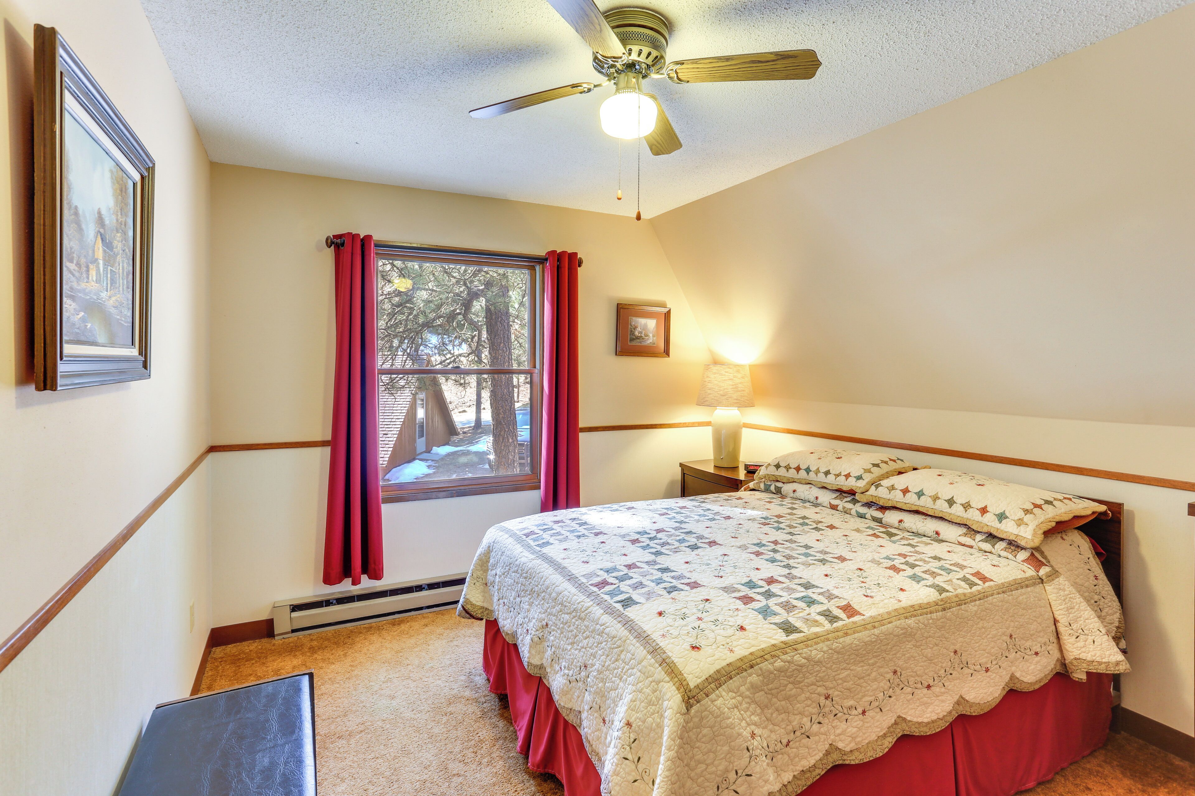Um quarto com uma cama, um ventilador de teto, uma janela com cortinas vermelhas e uma luminária em uma mesa de cabeceira.