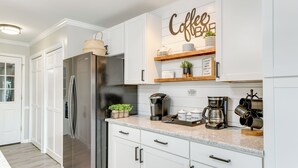 Uma cozinha moderna com armários brancos, geladeira de aço inoxidável, um espaço para café com prateleiras e cafeteira.