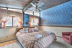 Quarto com cama, ventilador de teto, janela com cortinas, parede com papel de parede floral e uma mesinha de cabeceira com abajur.