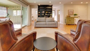 Uma sala de estar espaçosa com um sofá cinzento, poltronas de couro e uma mesa de centro. Uma estante grande e um armário verde são visíveis ao fundo.