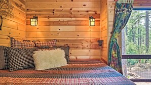 Quarto de cabana de madeira com colcha xadrez, duas lanternas fixadas na parede e uma janela com cortina.