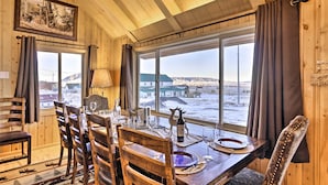 Interior em madeira com uma mesa de jantar posta para quatro pessoas, um quadro de animais emoldurado e vista para uma paisagem nevada através de grandes janelas.