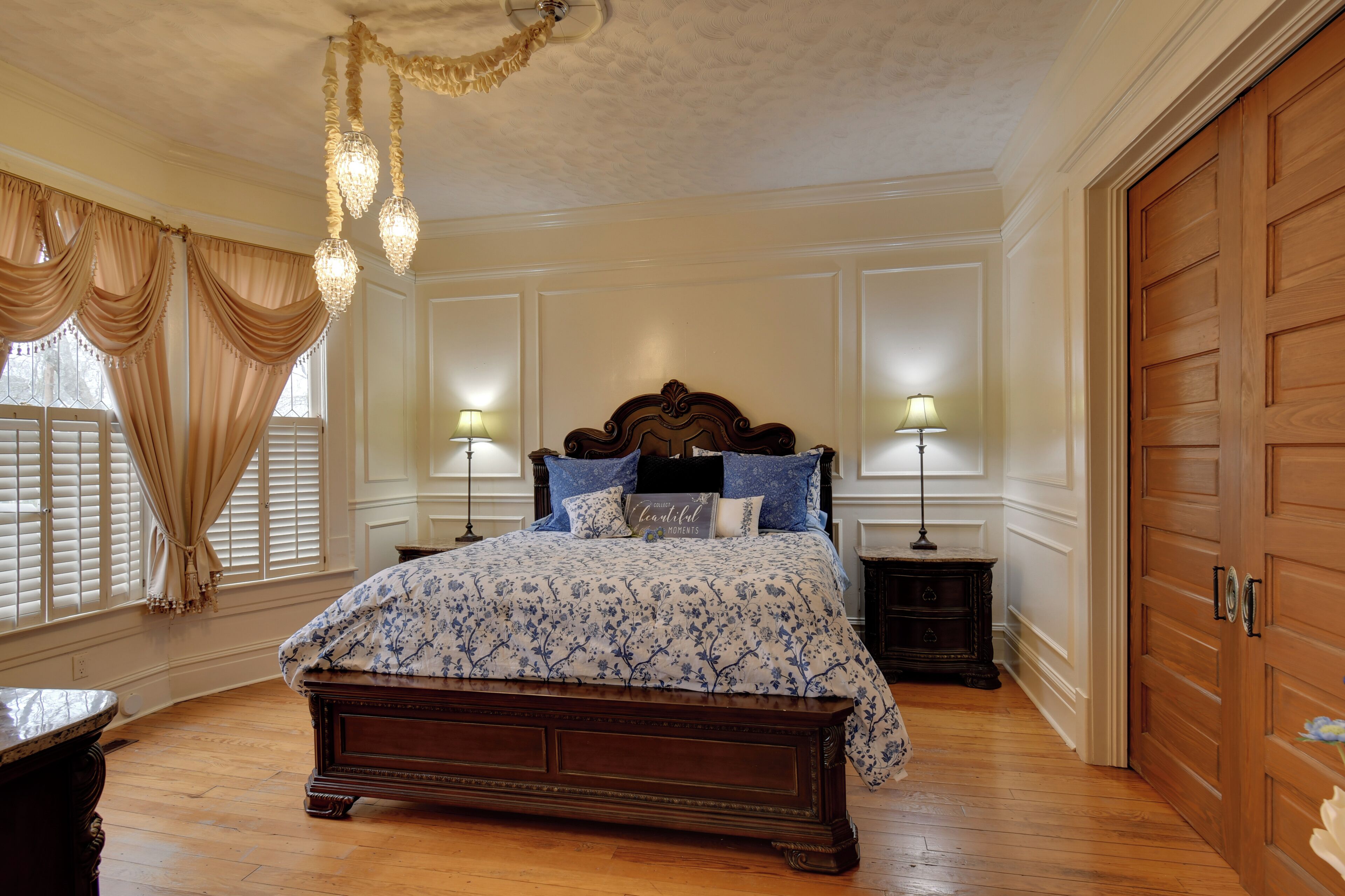 Quarto com cama de madeira, duas mesas de cabeceira com luminárias, um lustre e uma grande porta de madeira.