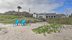 Uma casa de praia com duas cadeiras azuis e uma área arenosa com plantas verdes.