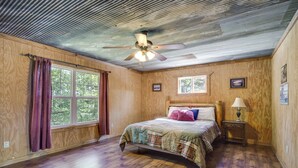 Quarto com teto de madeira, cama com colcha xadrez, mesa de cabeceira com abajur e janela com cortinas.