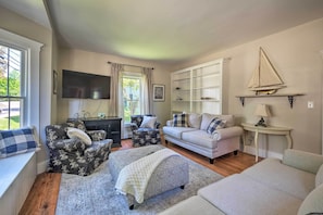 Uma sala de estar com sofá, poltrona, banquinho, TV, estante de livros e decoração náutica com um veleiro.