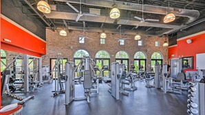 Uma academia ampla com diversos equipamentos de exercício, janelas grandes e um fundo de parede de tijolos.