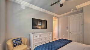 Quarto com cama, cômoda, cadeira, ventilador de teto e TV fixada na parede.