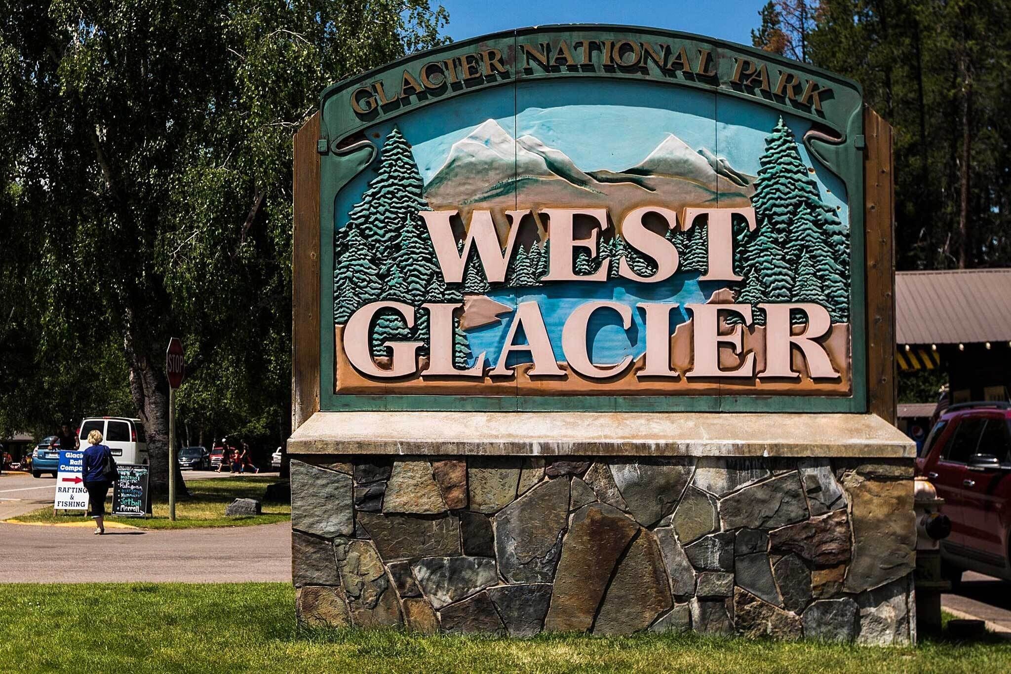 Placa indicando a região de West Glacier, no Parque Nacional Glacier, com paisagem montanhosa e árvores.