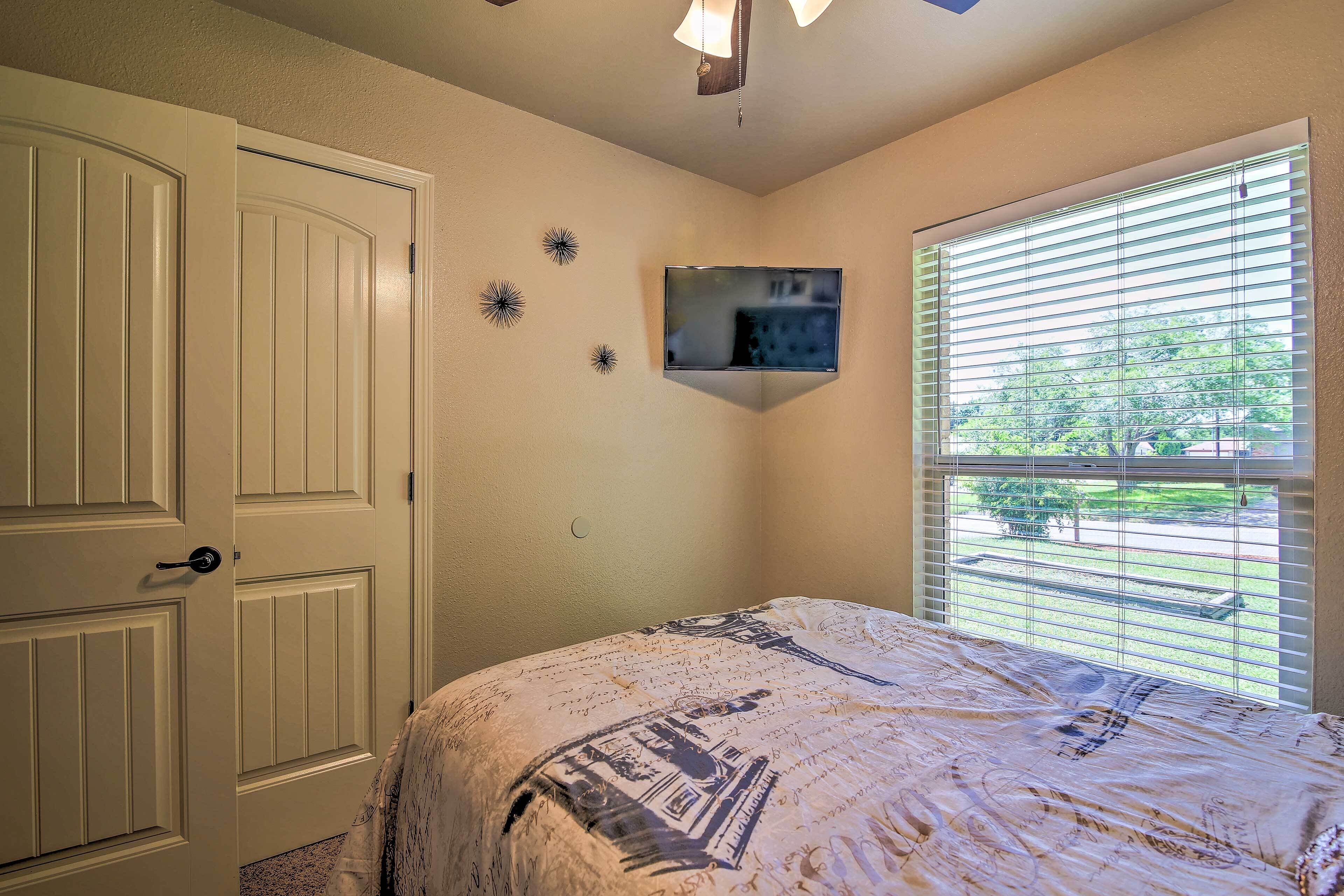 Quarto com cama, televisão presa na parede, janela com persianas e porta.