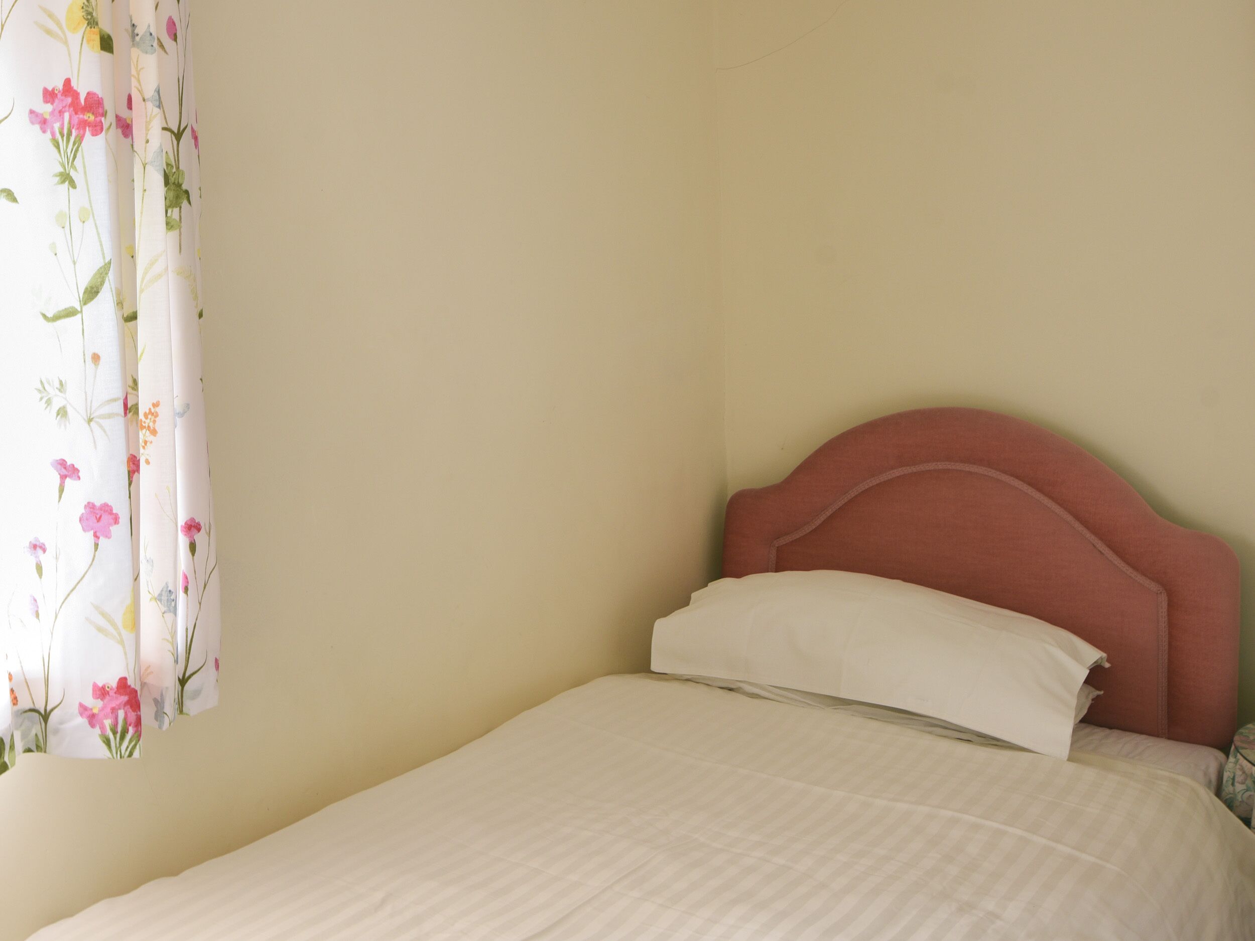 Uma cama de solteiro com cabeceira, uma parede lisa e uma janela com cortinas florais.
