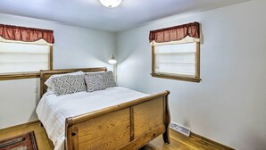 Quarto com cama de madeira, duas janelas com persianas, uma luminária no teto e um respiradouro no chão.