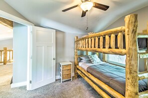 Um quarto com uma cama beliche de madeira, um ventilador de teto, uma mesinha de cabeceira com cestos e uma porta que dá para outro cômodo.