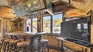 Uma área de bar com banquetas de madeira, uma televisão grande e uma janela de vitrais.