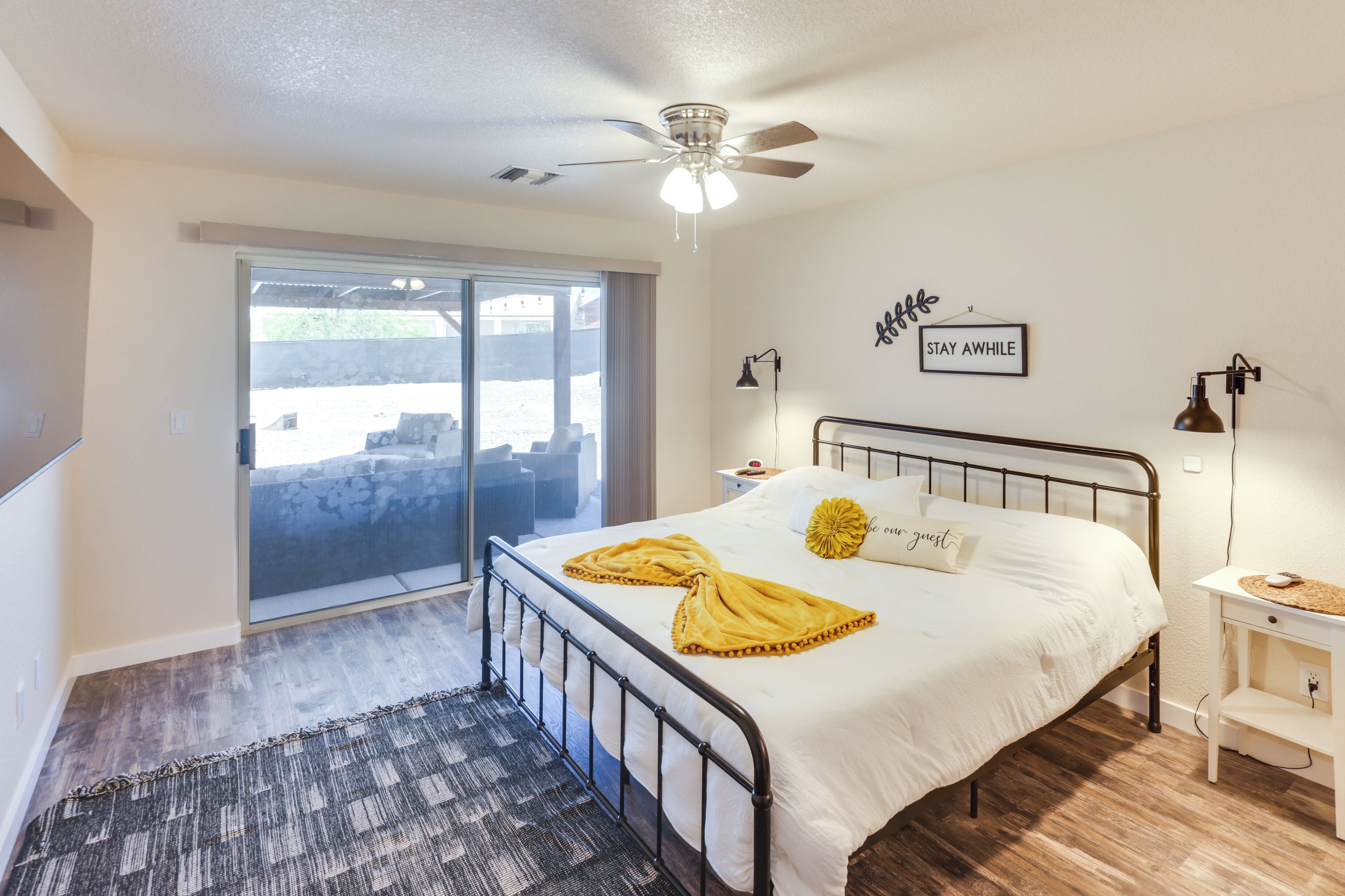 Quarto com cama, ventilador de teto, mesa de cabeceira, abajur, parede com um letreiro e vista através de uma porta deslizante.