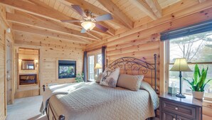 Quarto de cabana de madeira com uma cama, um ventilador de teto, mesa de cabeceira com abajur e janela com vista para árvores.