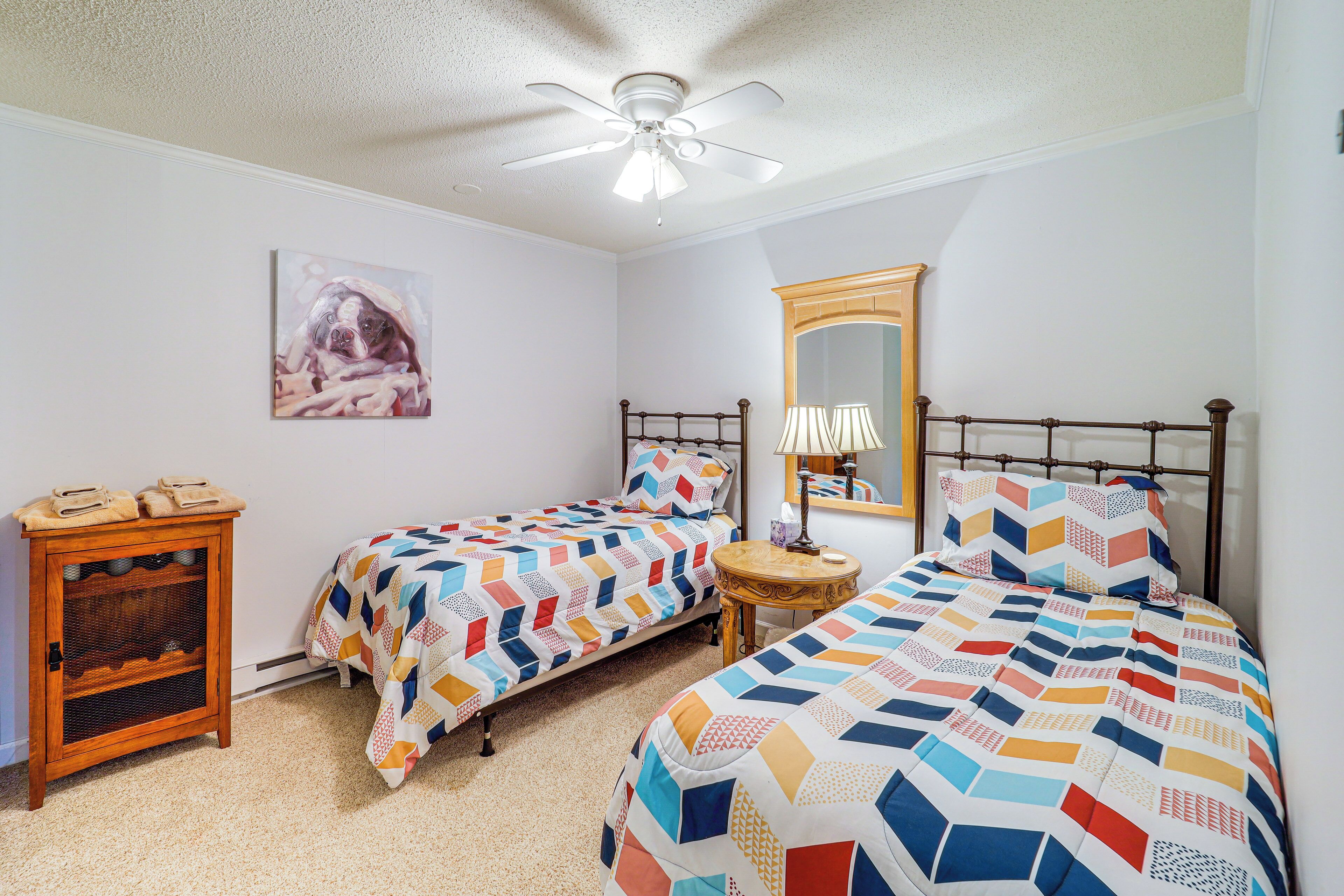 Quarto com duas camas de solteiro, uma mesinha de cabeceira de madeira, um ventilador de teto, um espelho e um quadro na parede.
