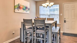 Uma sala de jantar com mesa e cadeiras de madeira, um candelabro, uma pintura de paisagem urbana e uma porta com tapete.