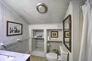Casa de banho com sanita, lavatório e zona de duche. Há prateleiras com toalhas e um quadro emoldurado na parede.