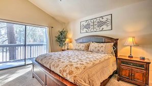Quarto com uma cama grande, mesa de cabeceira, luminária e vista para árvores por uma porta de correr.