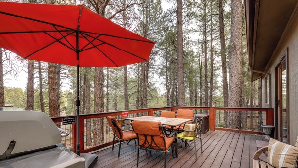 Um deck de madeira com mesa, cadeiras, churrasqueira e um guarda-sol vermelho.