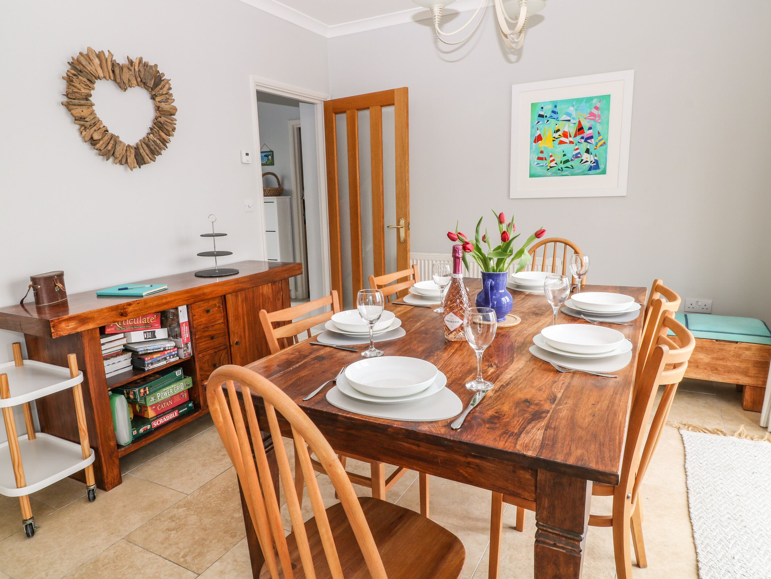 Uma sala de jantar com uma mesa de madeira posta para quatro pessoas, rodeada por cadeiras de madeira. Uma estante com jogos de tabuleiro fica à esquerda.