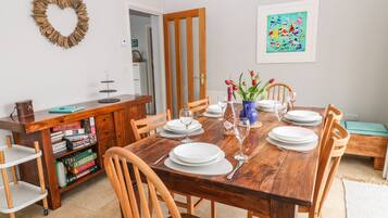 Uma sala de jantar com uma mesa de madeira posta para quatro pessoas, rodeada por cadeiras de madeira. Uma estante com jogos de tabuleiro fica à esquerda.
