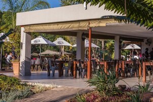 Um café ao ar livre com mesas e cadeiras sob um toldo, rodeado por vegetação.
