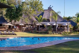 Uma piscina com espreguiçadeiras e guarda-sóis, cercada por edifícios com telhados de palha e áreas de estar ao ar livre.