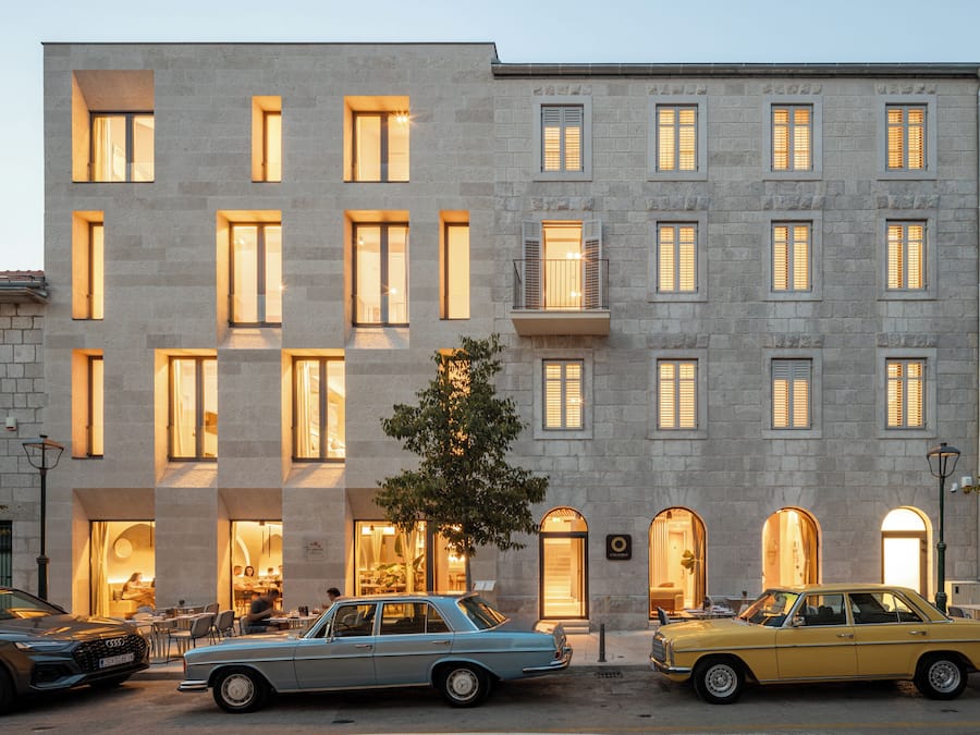 Um edifício de vários andares com fachada de pedra, várias janelas e varandas. Carros estão estacionados em frente e é possível ver um restaurante com mesas ao ar livre através das janelas.