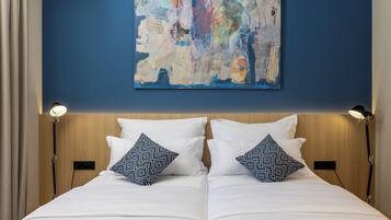 Uma cama de casal com roupa de cama branca e almofadas estampadas, um quadro abstrato azul na parede e duas luminárias de cabeceira.