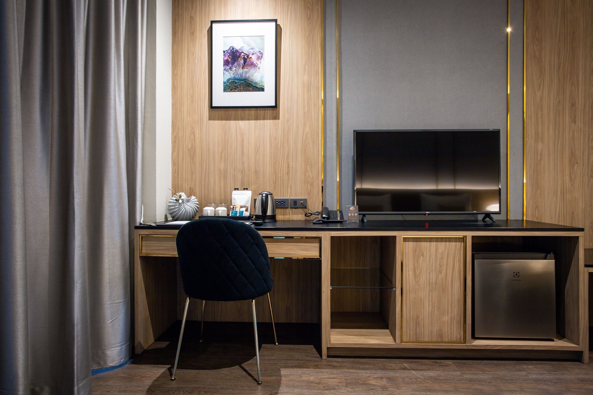 Quarto de hotel moderno com escrivaninha de madeira, TV de tela plana, pequena geladeira e uma cadeira.