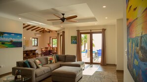 Uma sala de estar espaçosa com um sofá seccional, uma mesa de centro de vidro e um ventilador de teto. É possível ver uma área de jantar com mesa e cadeiras através de uma porta de vidro.