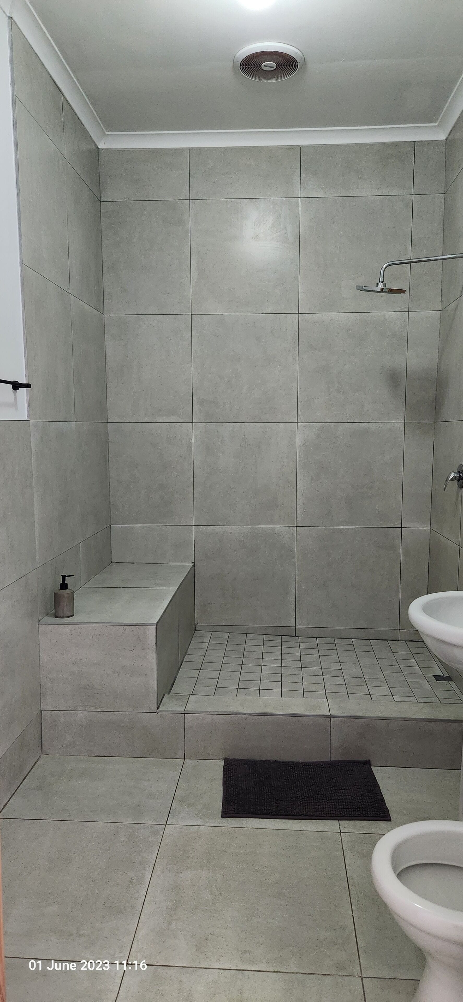 Uma casa de banho moderna com azulejos cinzentos, zona de duche, sanita e um pequeno armário com uma garrafa.