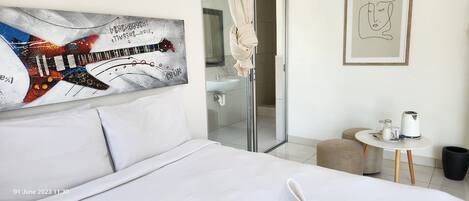 Quarto de hotel com cama, pintura de uma guitarra, banheiro com chuveiro, pequena mesa com chaleira e quadro na parede.