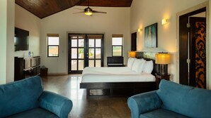 Quarto de hotel com uma cama grande, dois sofás azuis, um ventilador de teto, uma televisão e vista pela porta.
