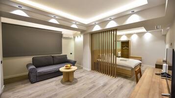Quarto de hotel moderno com sofá, mesa redonda pequena com fruteira, estrutura de cama de madeira, escrivaninha de madeira e televisão.