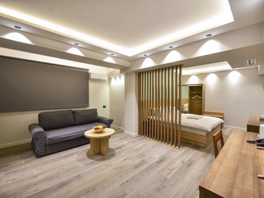 Quarto de hotel moderno com sofá, mesa redonda pequena com fruteira, estrutura de cama de madeira, escrivaninha de madeira e televisão.