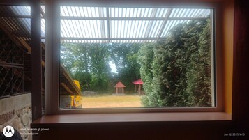 Visão pela janela com telhado de metal, um veículo amarelo e uma construção com telhado vermelho numa área gramada.