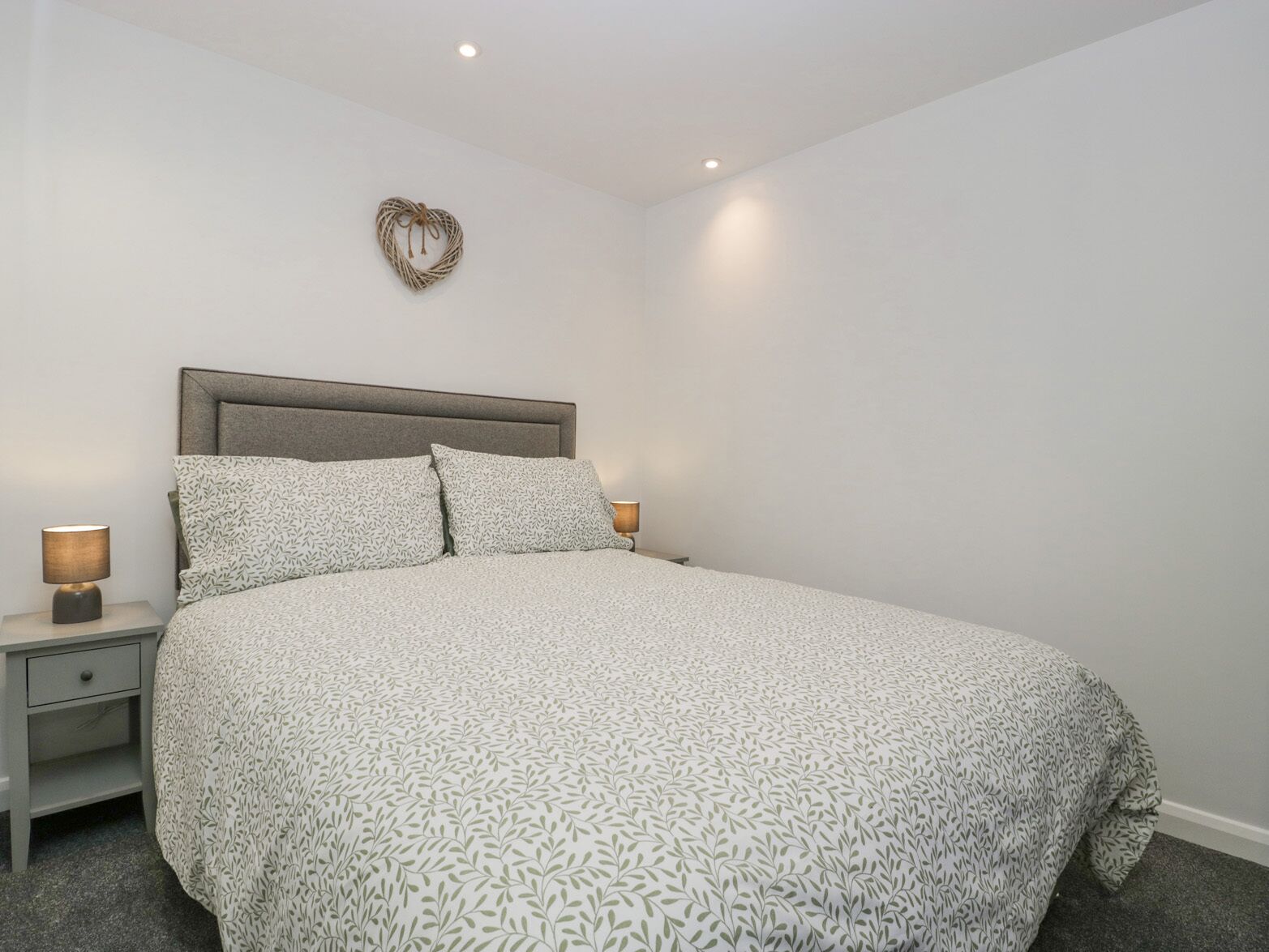 Um quarto com uma cama grande, duas mesas de cabeceira com abajures, um enfeite em formato de coração na parede e um teto branco liso.
