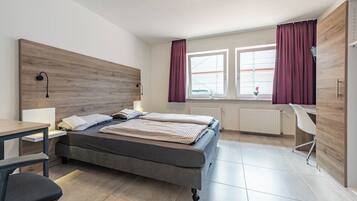 Quarto de hotel moderno com uma cama, uma escrivaninha e uma cadeira. Possui duas janelas com cortinas, um radiador e cabeceira de madeira.