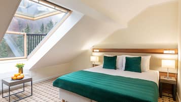 Quarto com claraboia, cama com roupa de cama verde, mesa de cabeceira com abajur e pequena mesa com frutas.